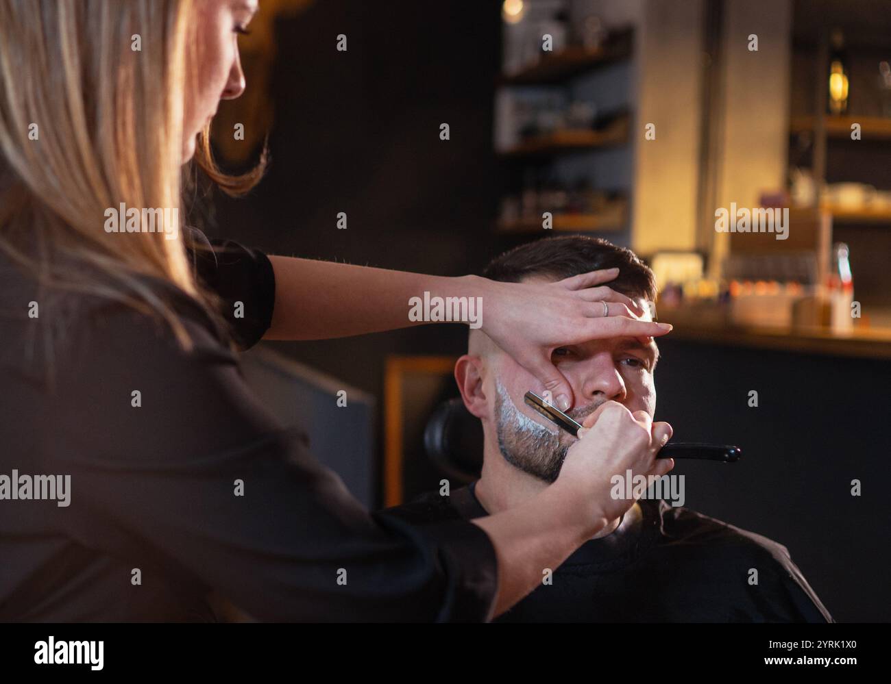 Long-haired young female Barber gently shaving bearded man face bristle ...
