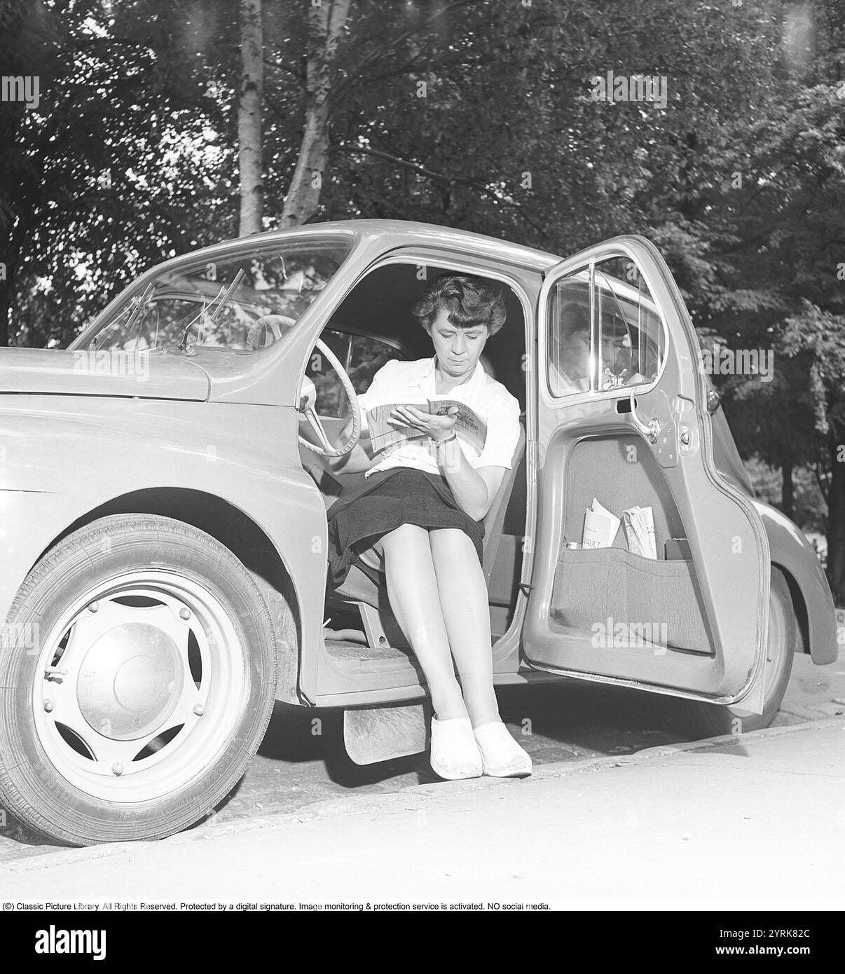 In the 1950s. A woman in the drivers seat of a small car reading a book ...