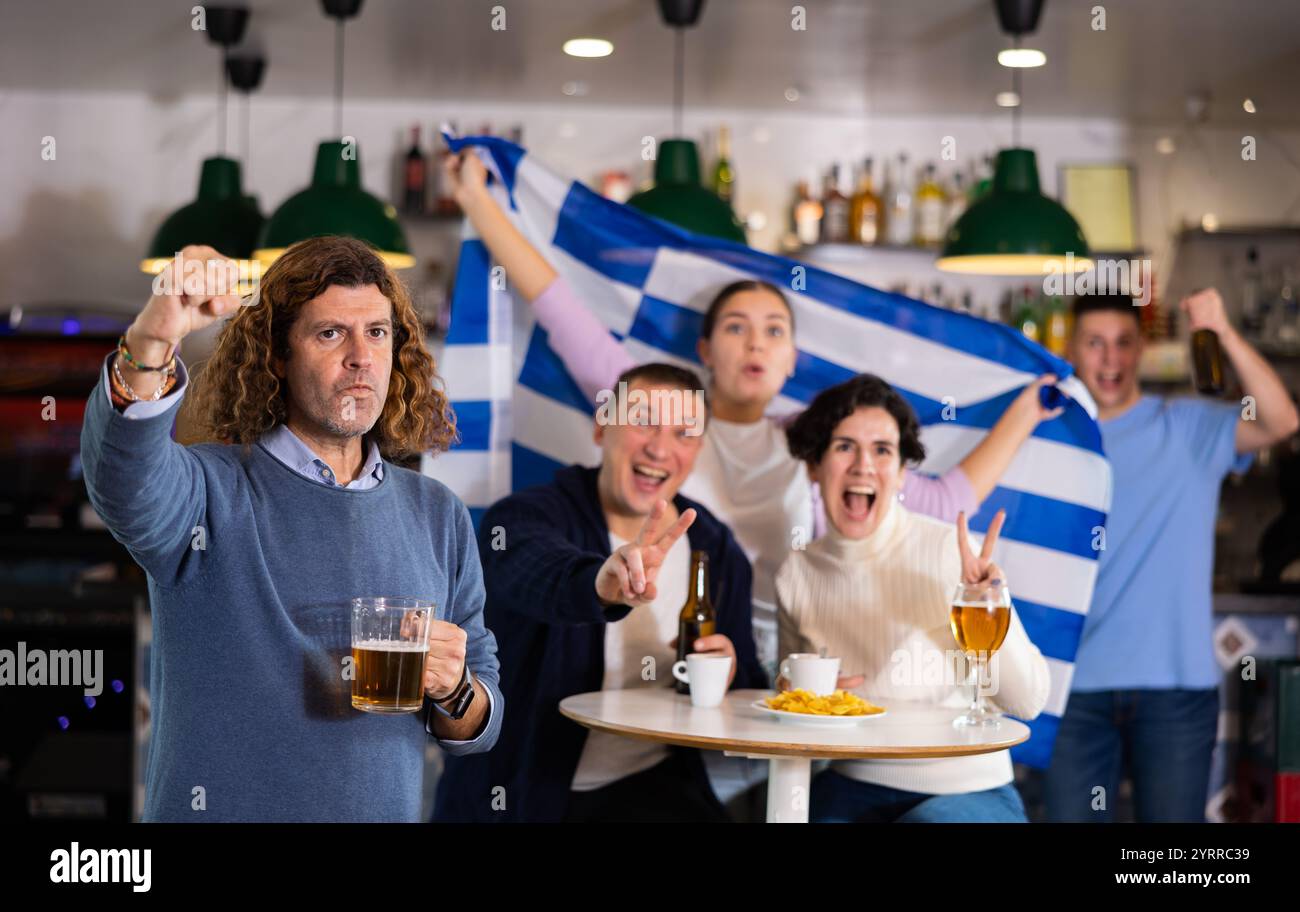 Company of excited young adult sports fans waving flag of Greece and ...