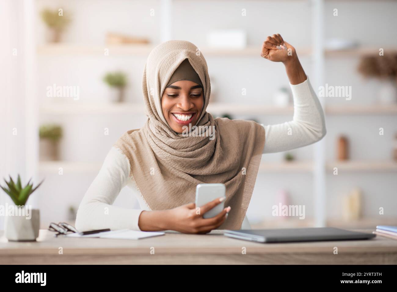 Overjoyed black muslim woman in hijab looking at smartphone and ...