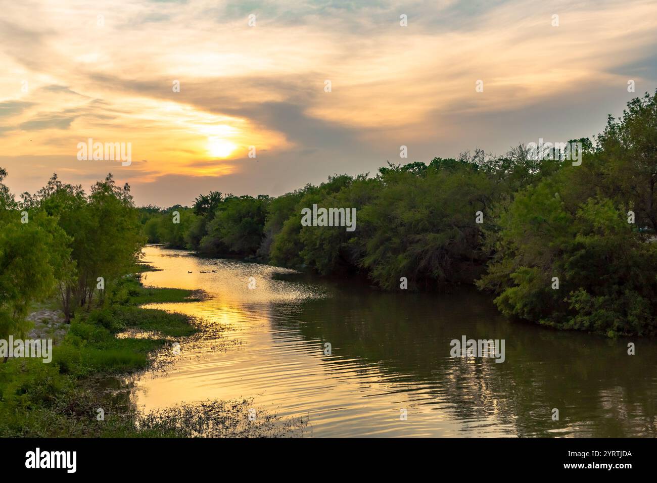 Dramatic golden sunset and clouds reflected along a treelined river ...