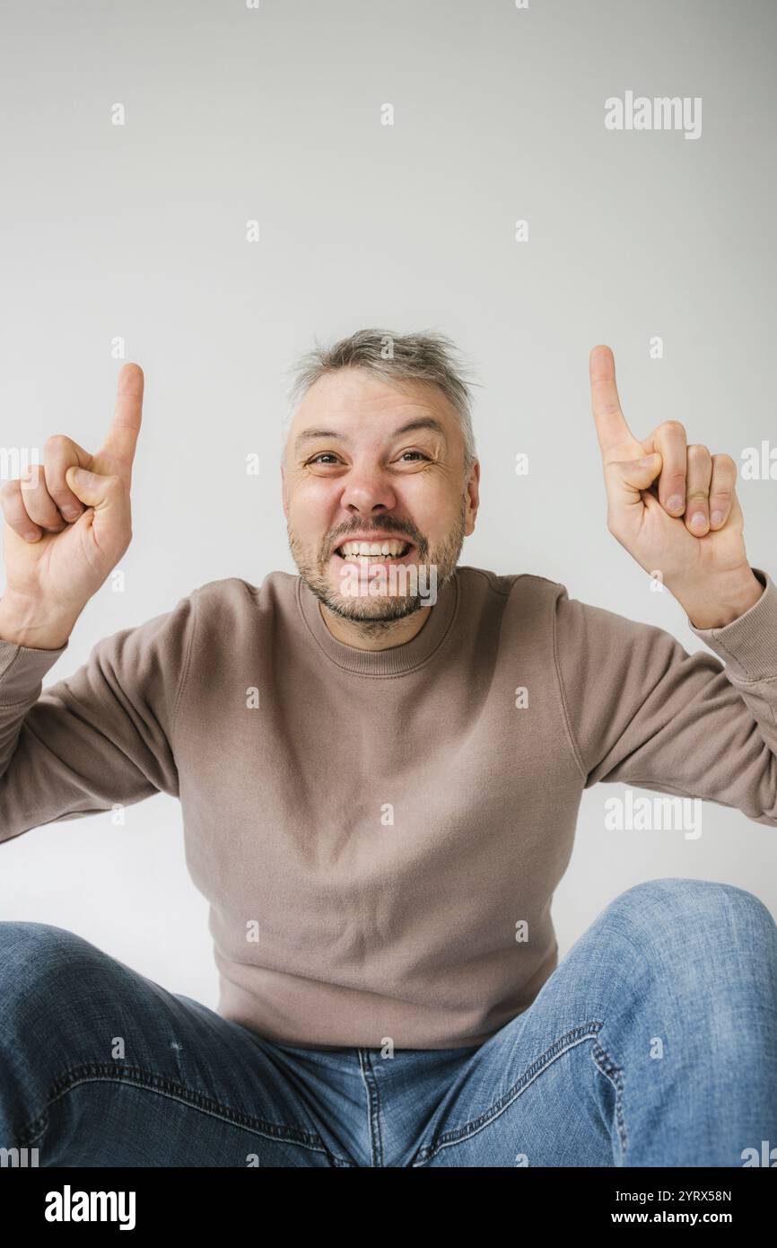 A playful portrait of a man sitting, pointing upwards with both hands ...