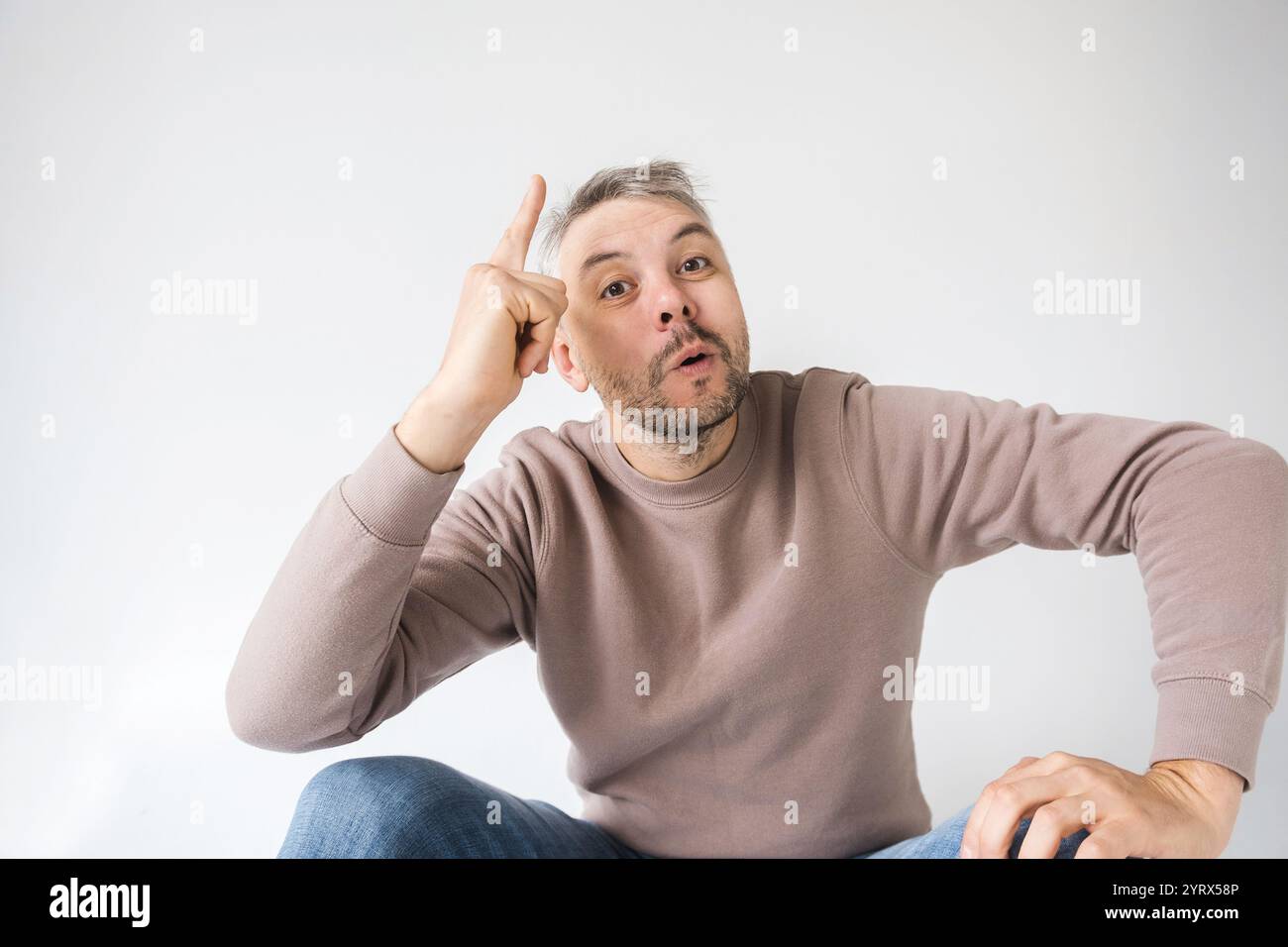 A man sits against a white background, pointing to his head with a ...