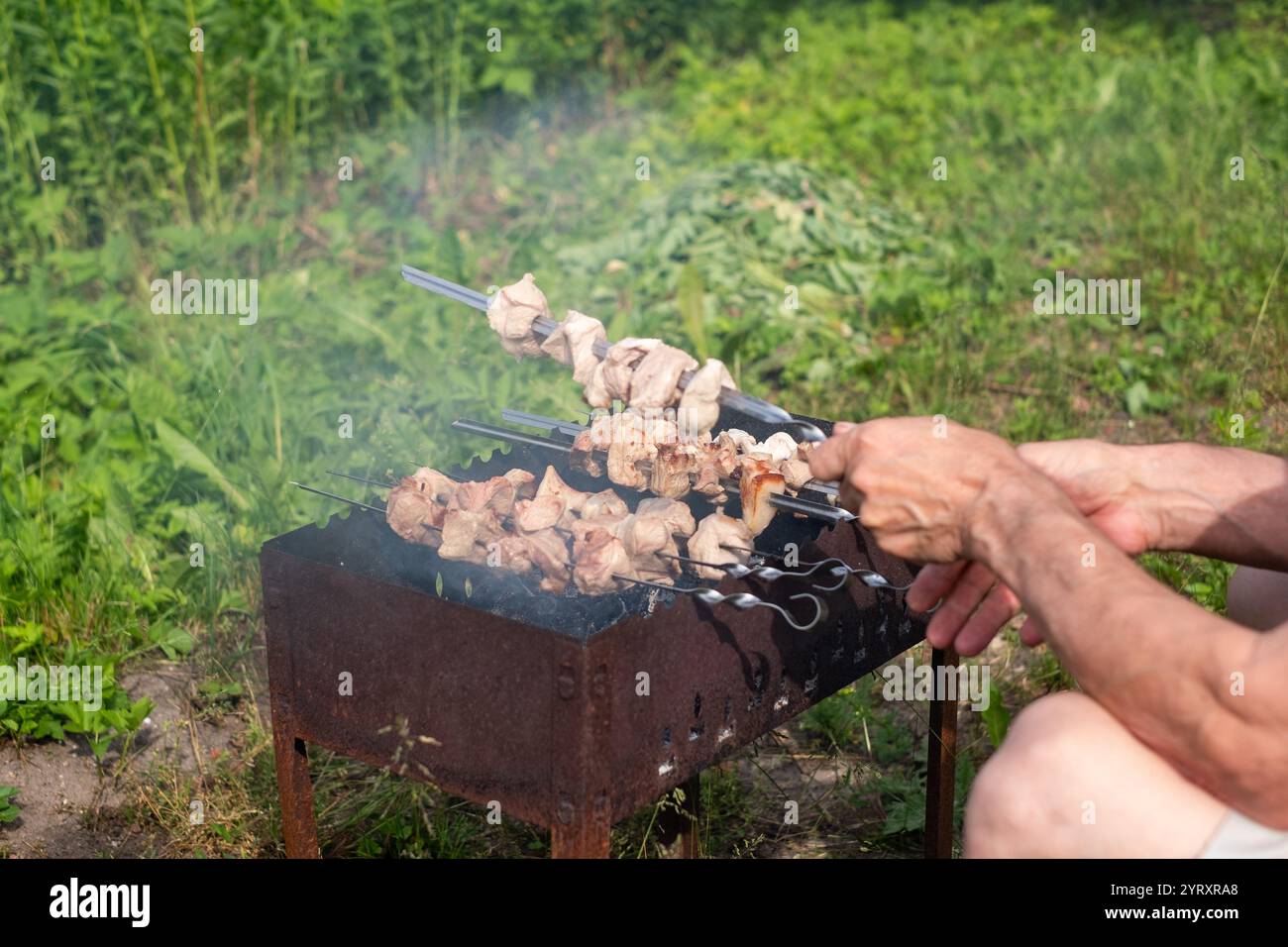 Skewers of meat are grilling over hot coals on a rusted barbecue. A ...