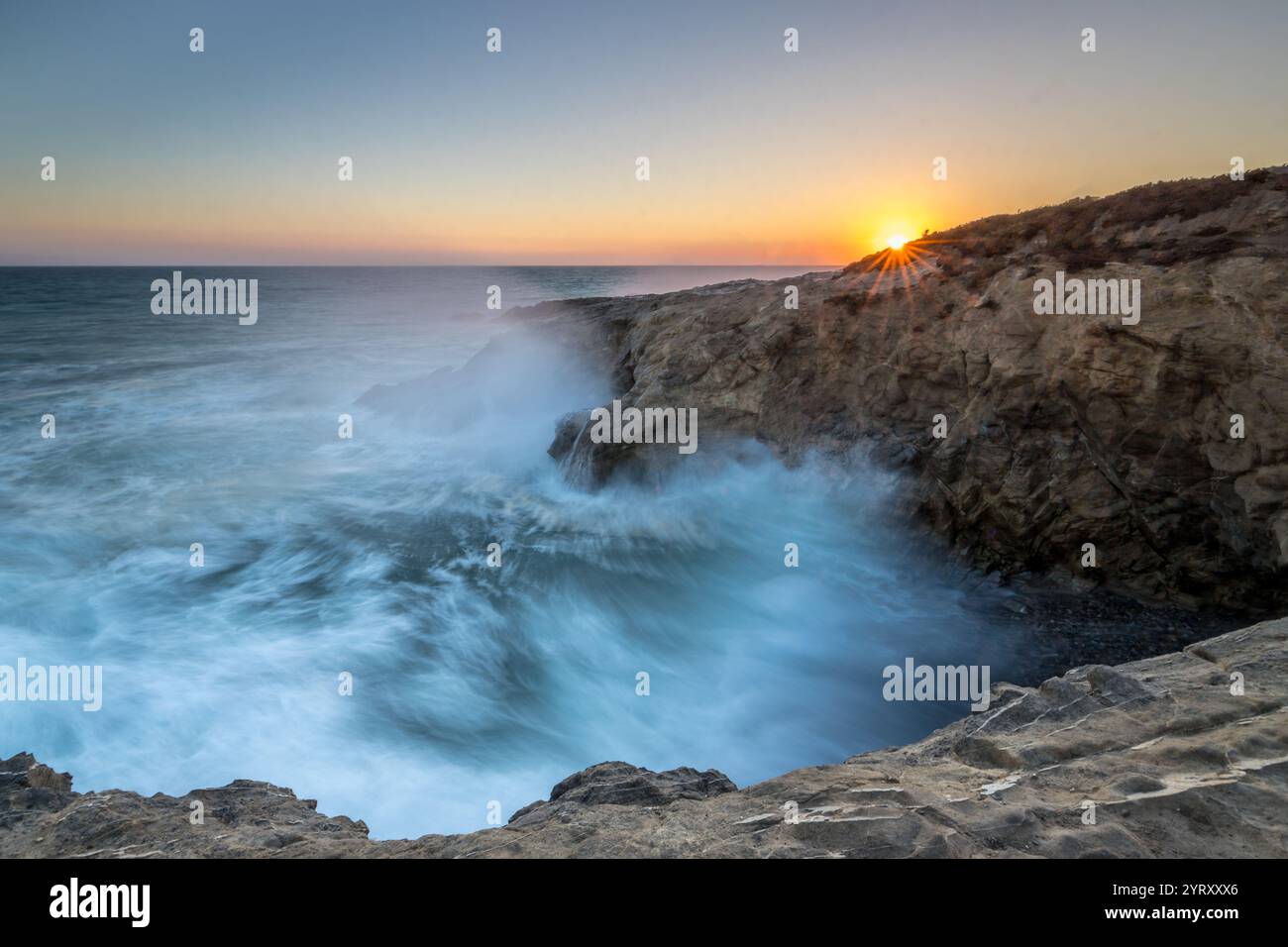 Waves crash against rugged cliffs as the sun sets, creating a ...