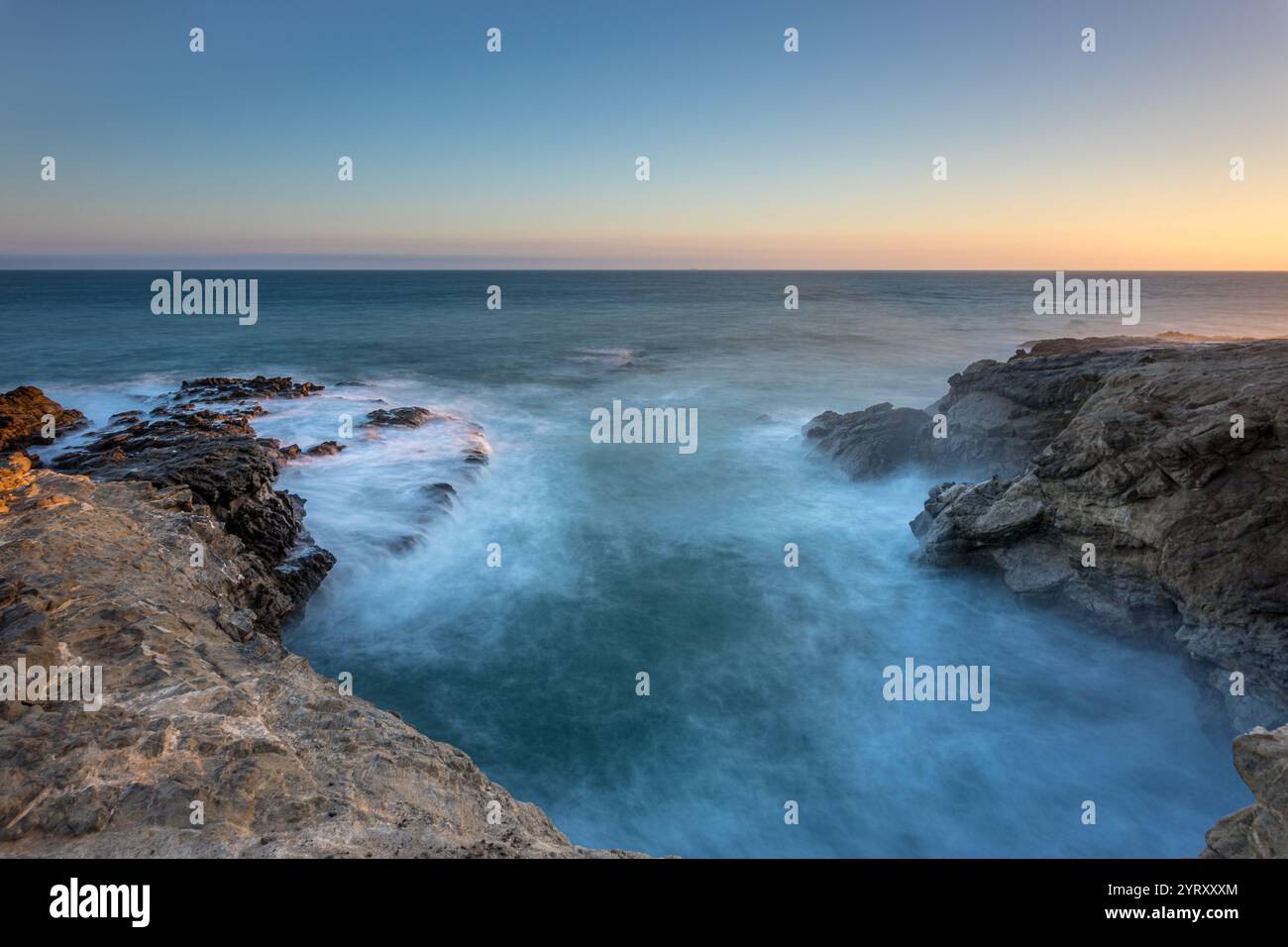 Waves gently crash against rocky cliffs as the sun sets, illuminating ...