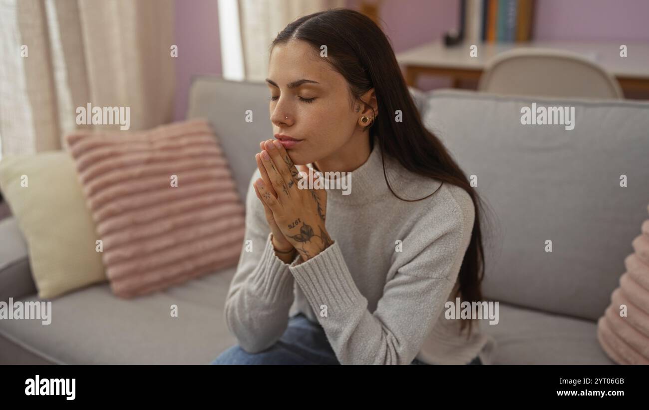 Woman praying indoors on a sofa with closed eyes in a cozy living room ...