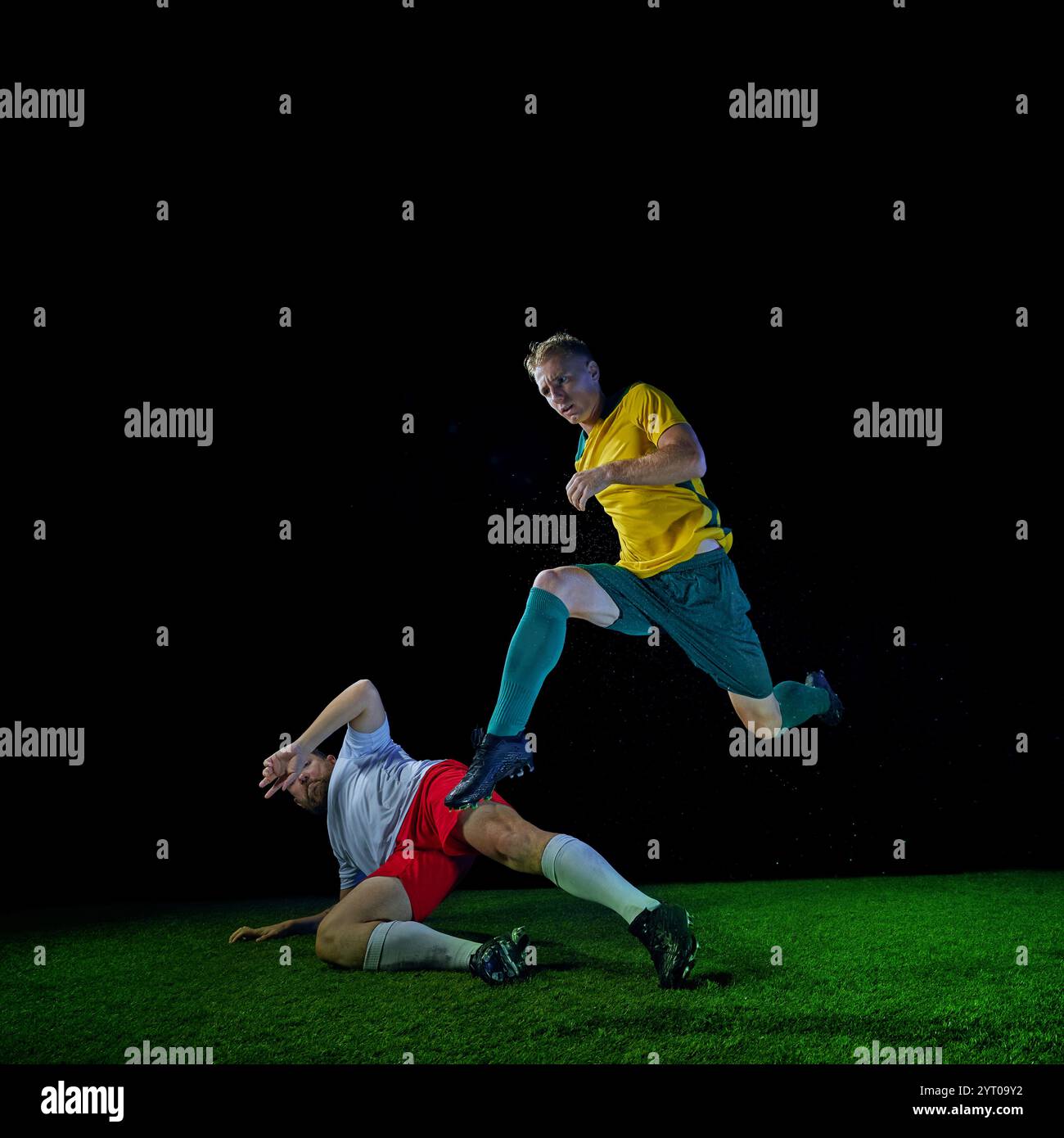 Player in yellow and green uniform jumps over fallen opponent on green ...