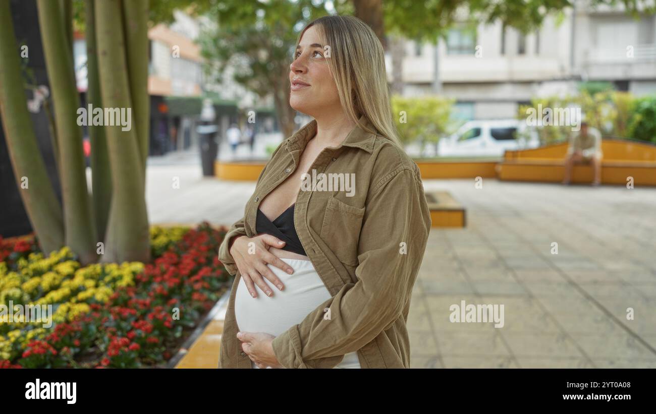 Pregnant woman with blonde hair stands outdoors in urban park, cradling ...
