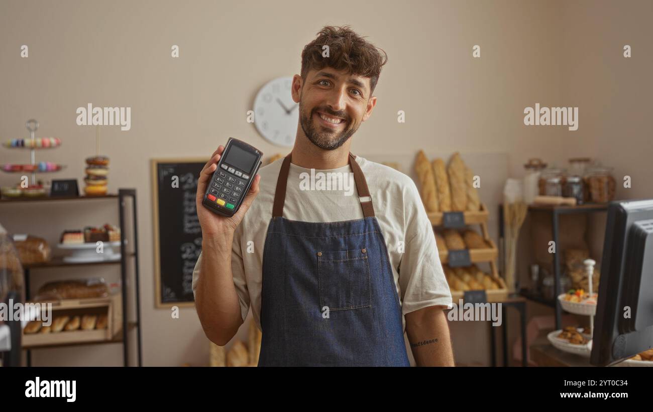 Young man in a bakery holding a card reader while wearing an apron and ...