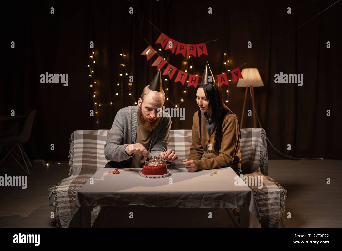 Man in party hat lighting candles on birthday cake for his wife sitting on sofa in the evening ...