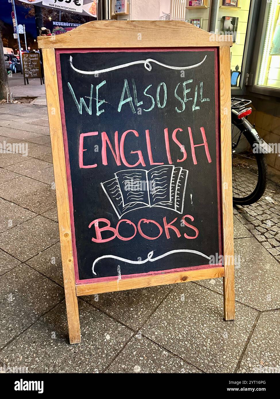 A wooden chalkboard on the sidewalk advertising "We also sell English ...