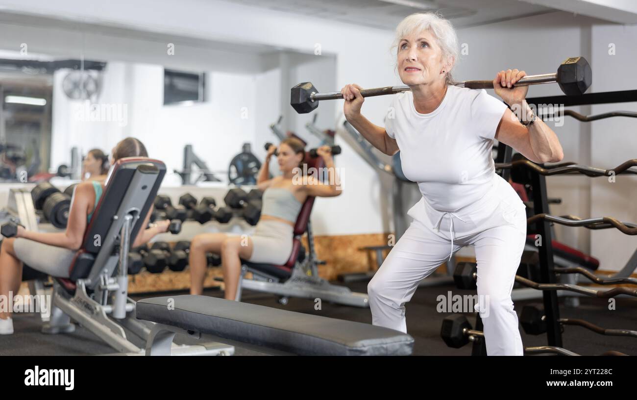 Senior woman trains muscles of arms and back using barbell and ...