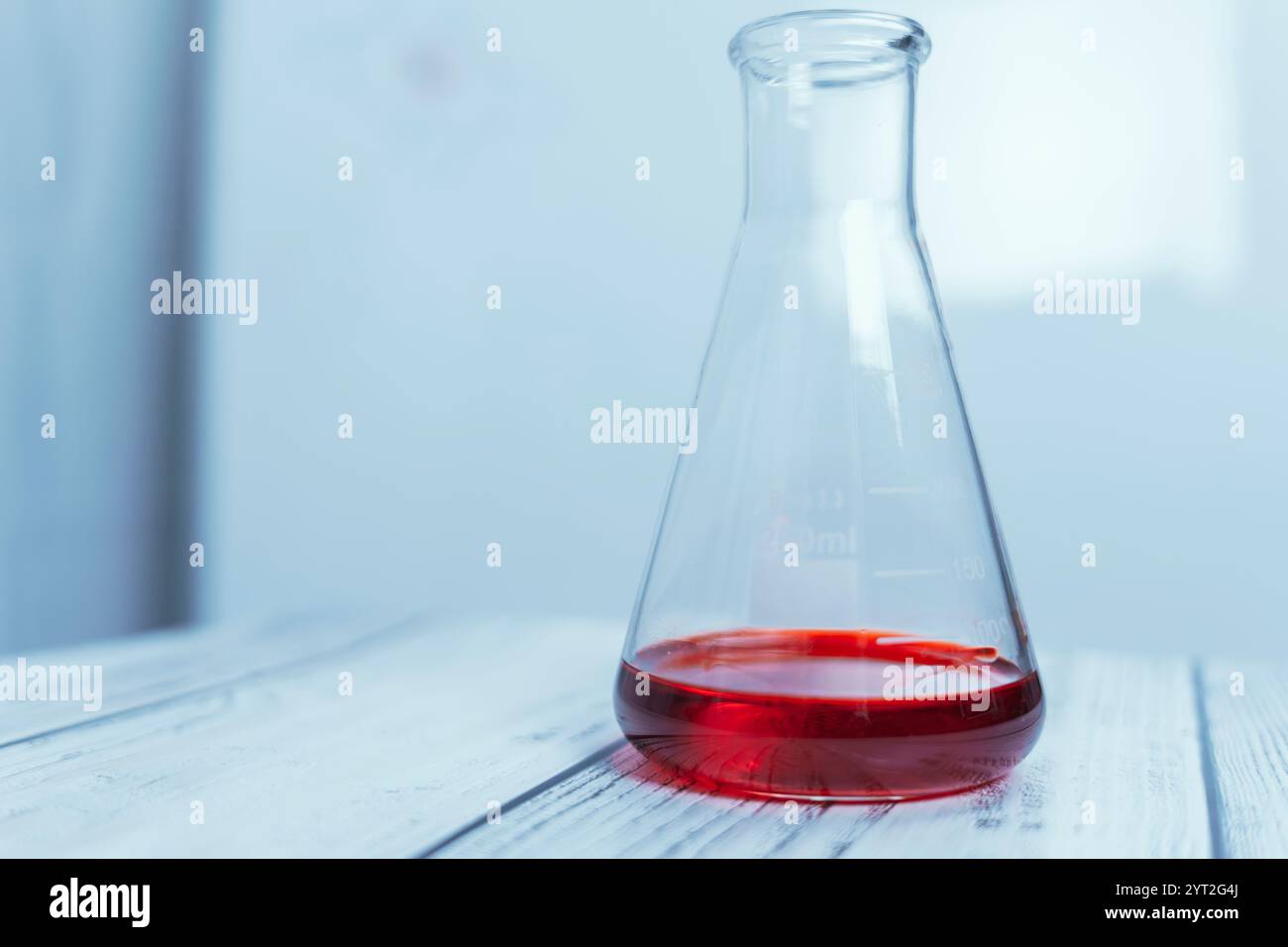 A glass Erlenmeyer flask with red liquid on a white surface. Scientific ...
