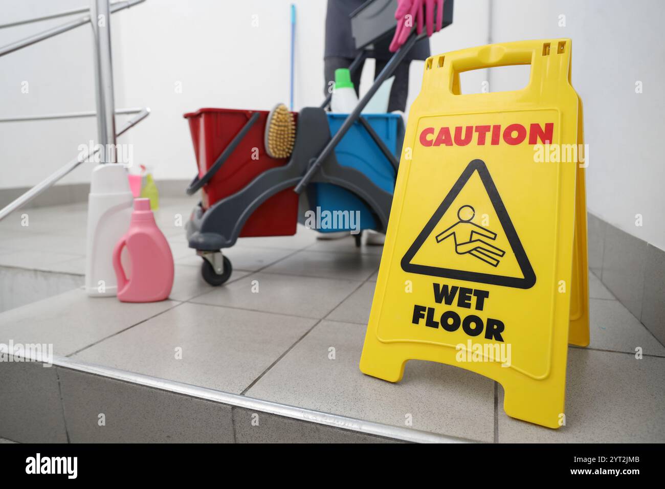 Caution sign on floor against female janitor with trolley of cleaning supplies in stairway ...