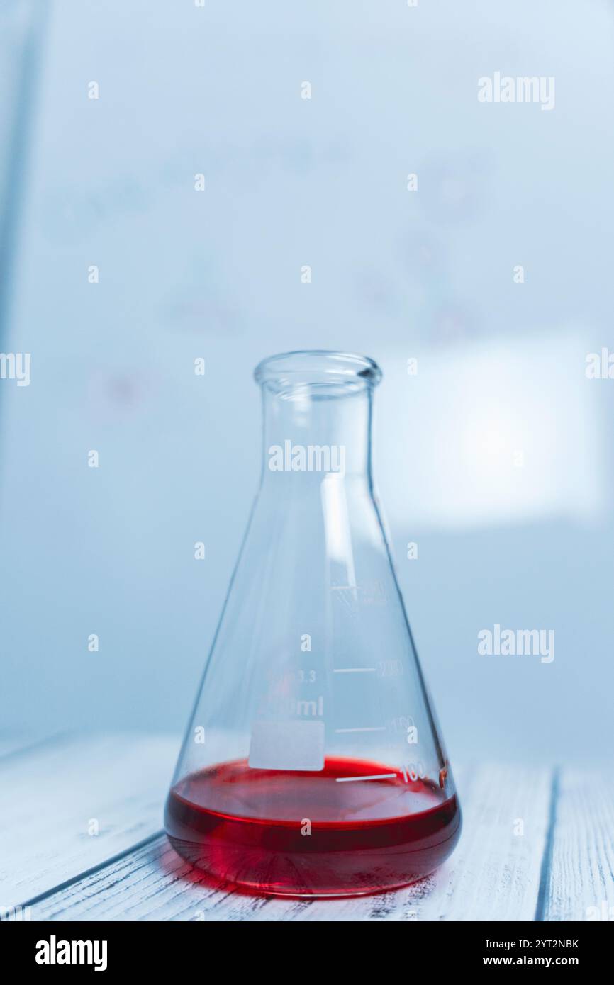 A glass Erlenmeyer flask with red liquid on a white surface. Scientific ...