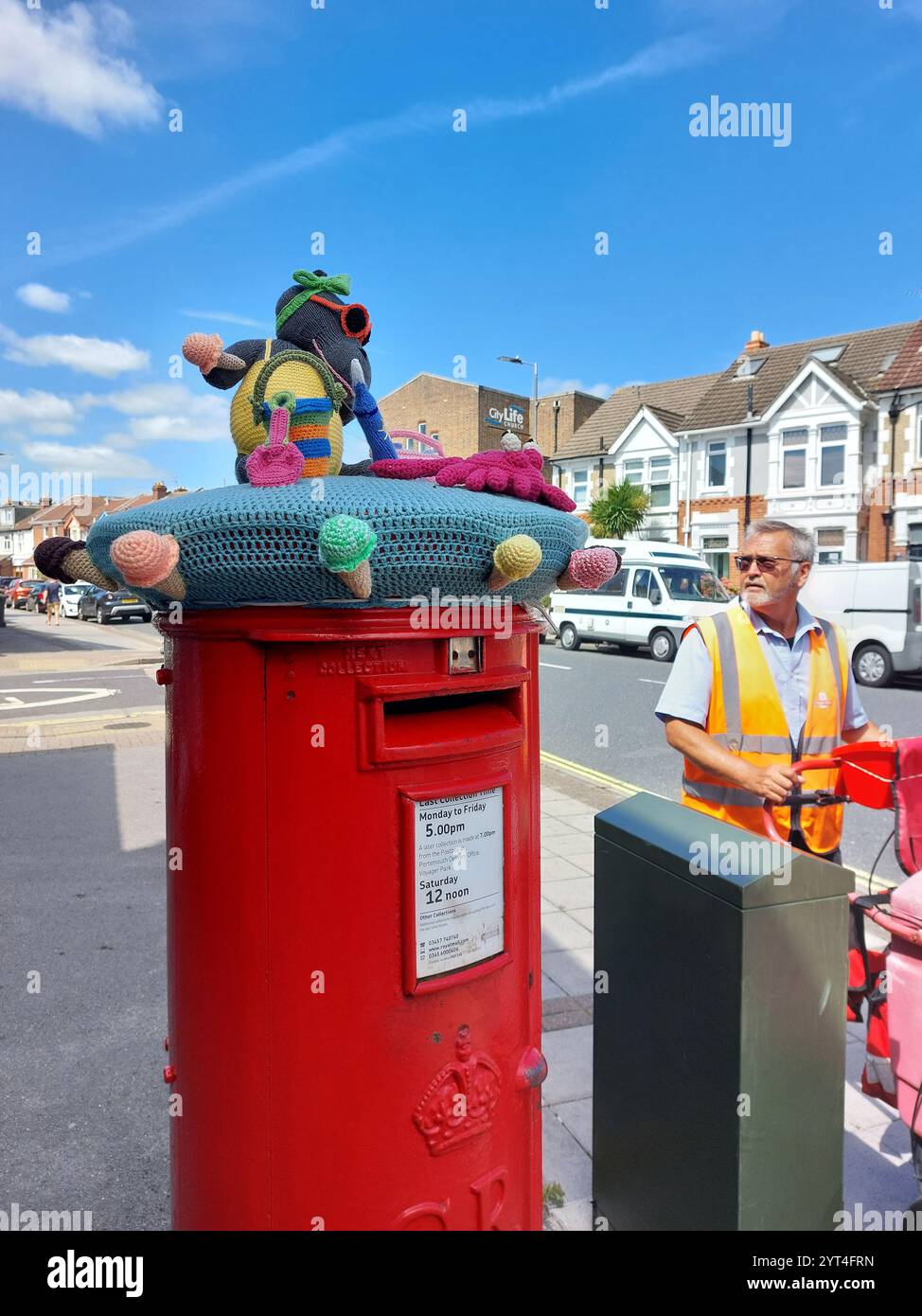 postman walking with trolley looking at post box with yarn bombed ...