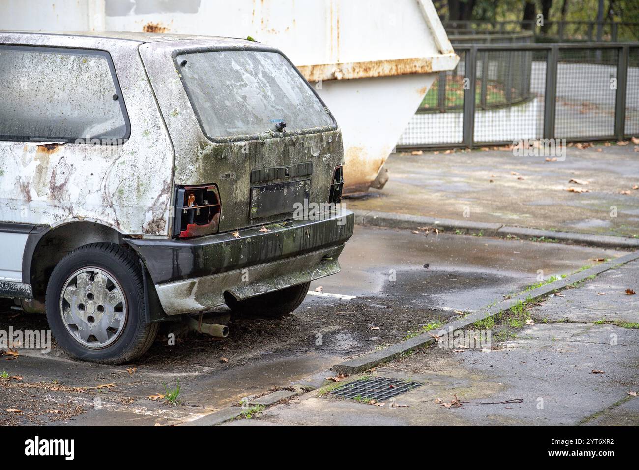 An abandoned, weathered car and rusted body sits in a neglected parking lot, covered in moss and ...