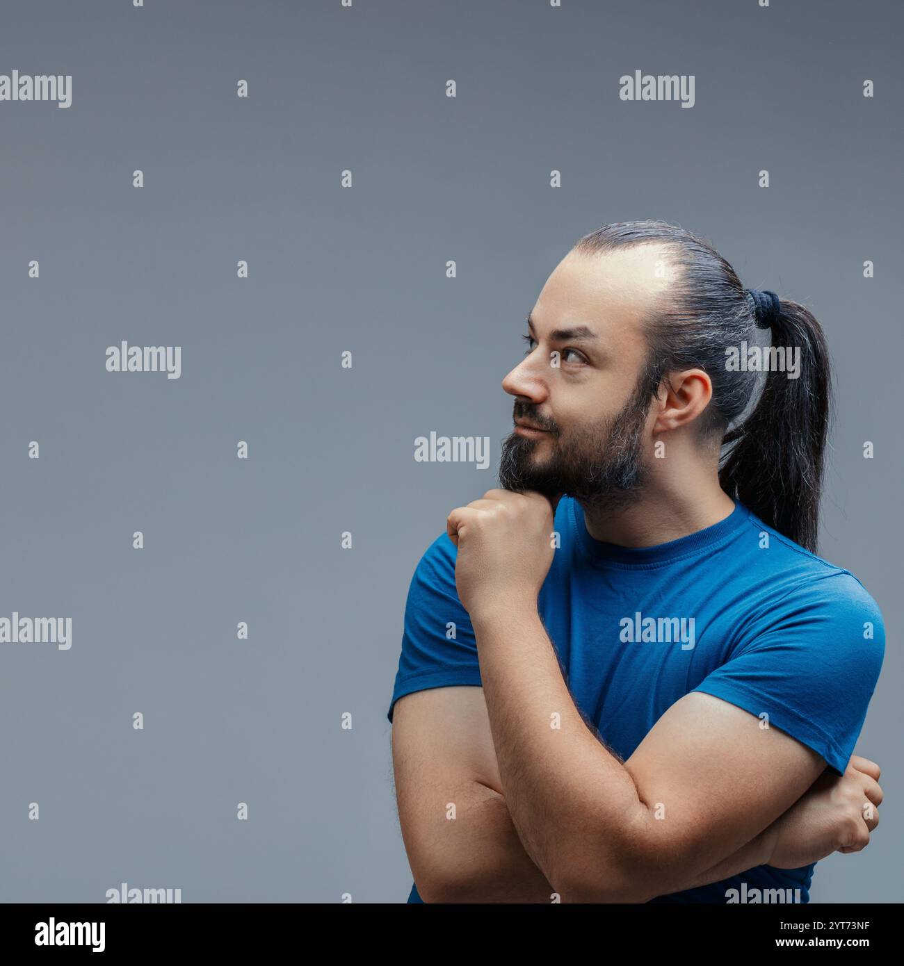 Thoughtful man with a ponytail and beard, wearing a blue t-shirt, holds ...