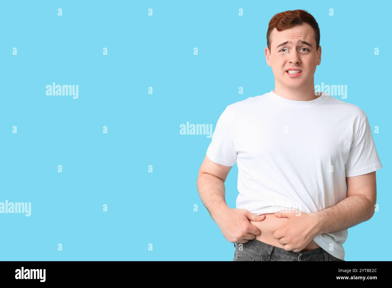 Young worried overweight man touching fat on his belly against blue ...