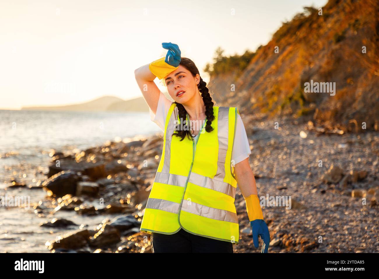 Tired young woman volunteer wearing vest and gloves wiping sweat from ...