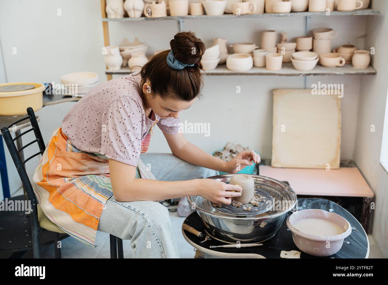 Young woman sculpting pottery on the wheel in a bright studio space ...