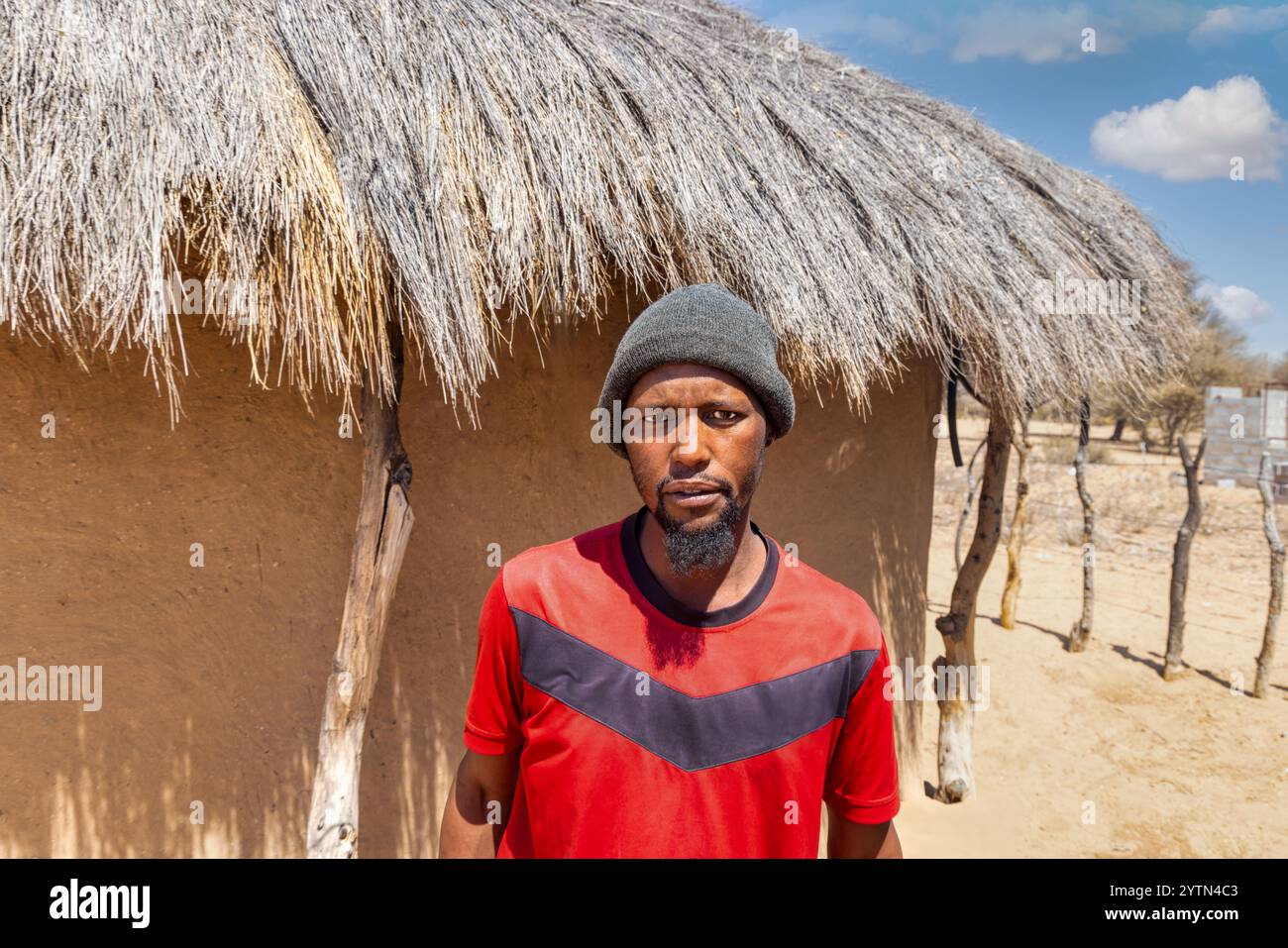 african village, young man with a beard and a beanie standing in front of a mud hut with ...
