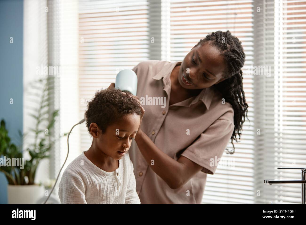 Caring young mother helping little son with drying hair in bathroom during daily hygiene routine ...