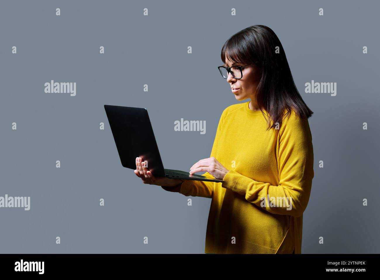 Serious middle aged woman holding laptop on grey studio background ...