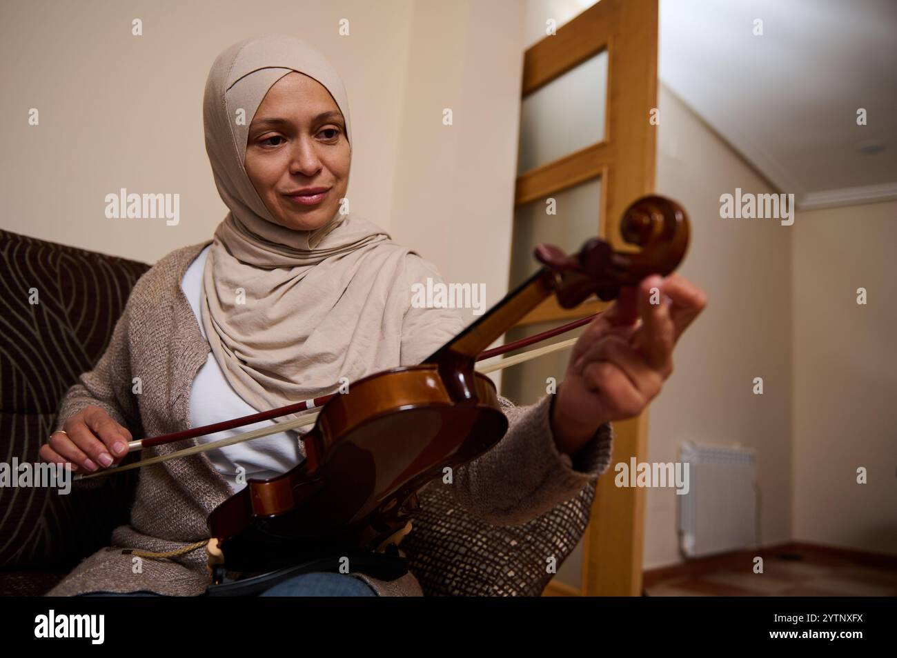 A woman wearing a hijab examines a violin at home, showcasing focus and dedication to music ...