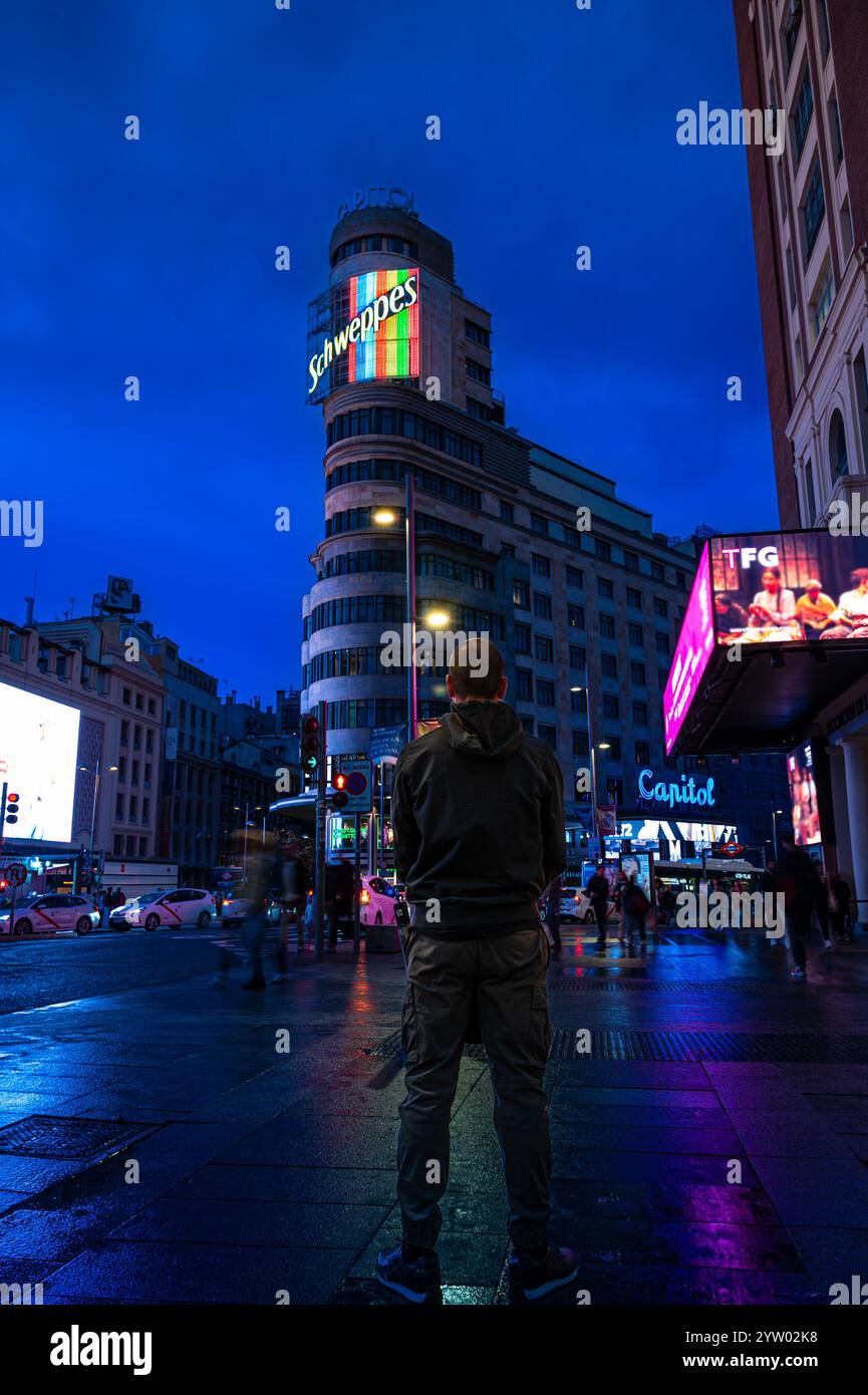 A man stands on a wet sidewalk in front of a building with a neon sign ...