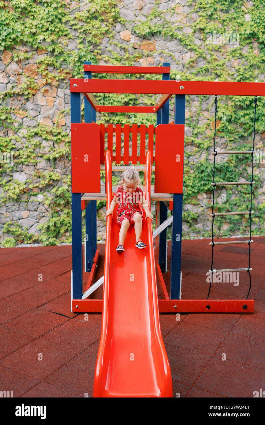 Little girl slides down a slide on a playground near a stone wall ...