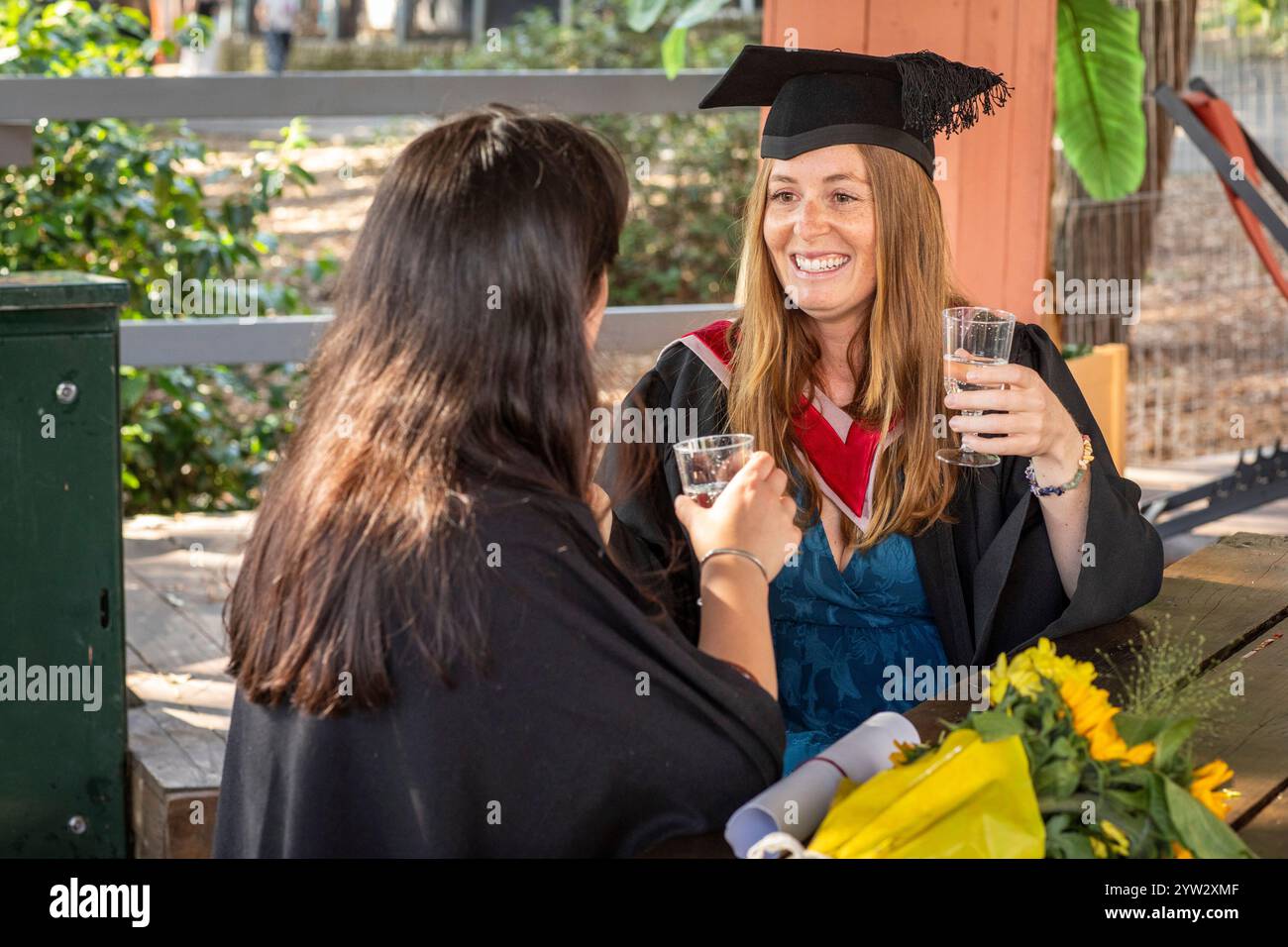 Smiling graduate in cap and gown celebrating with a friend over drinks ...