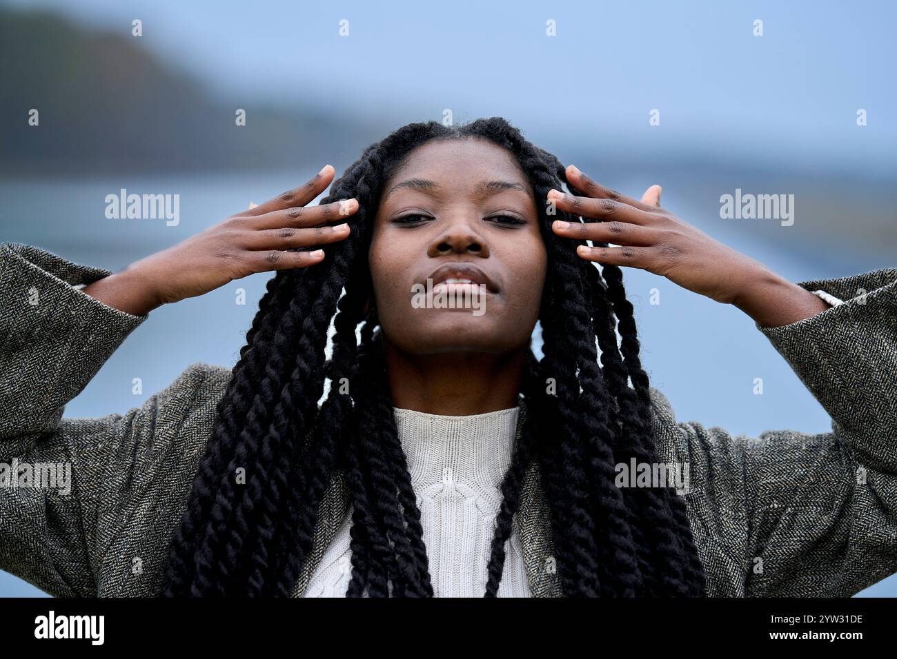 A serene young woman with long braids poses outdoors, her hands ...