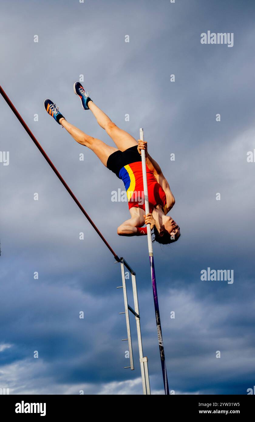 Female pole vaulter in mid-air over the bar against a cloudy sky ...