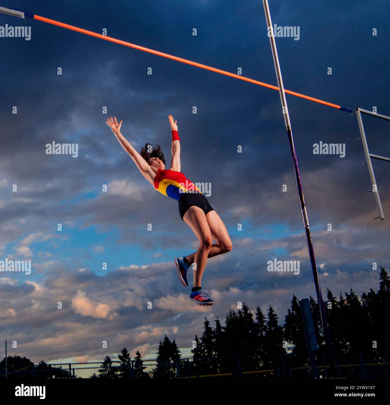 Female athlete in mid-air attempting a high jump over a bar at sunset ...