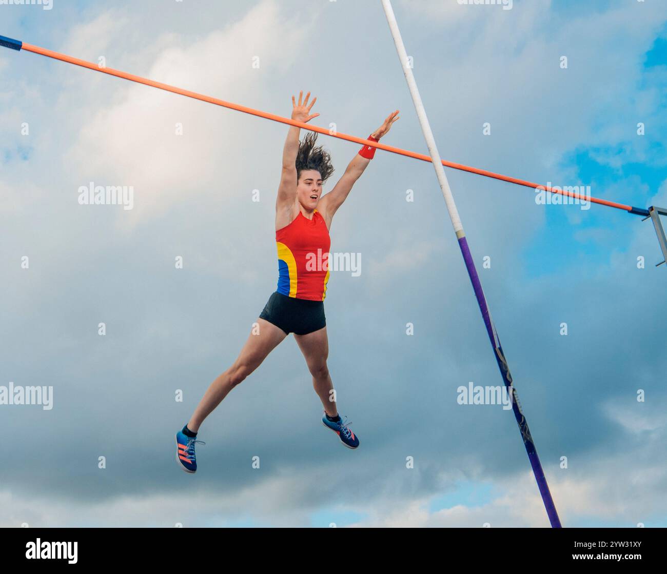 Female pole vaulter in mid-air clearing the bar against a cloudy sky ...