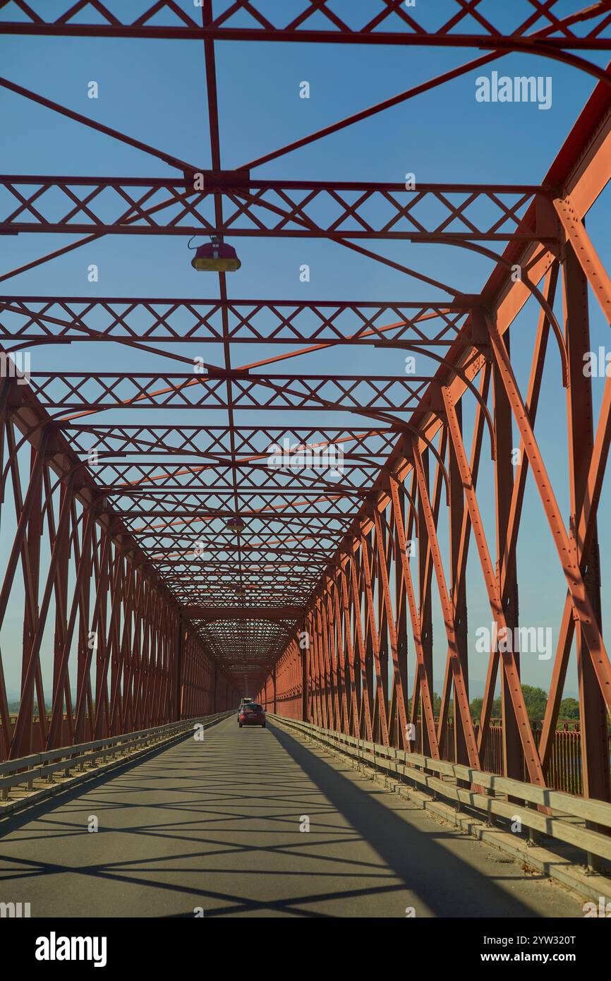 Red steel truss bridge under a clear blue sky with a single car driving ...