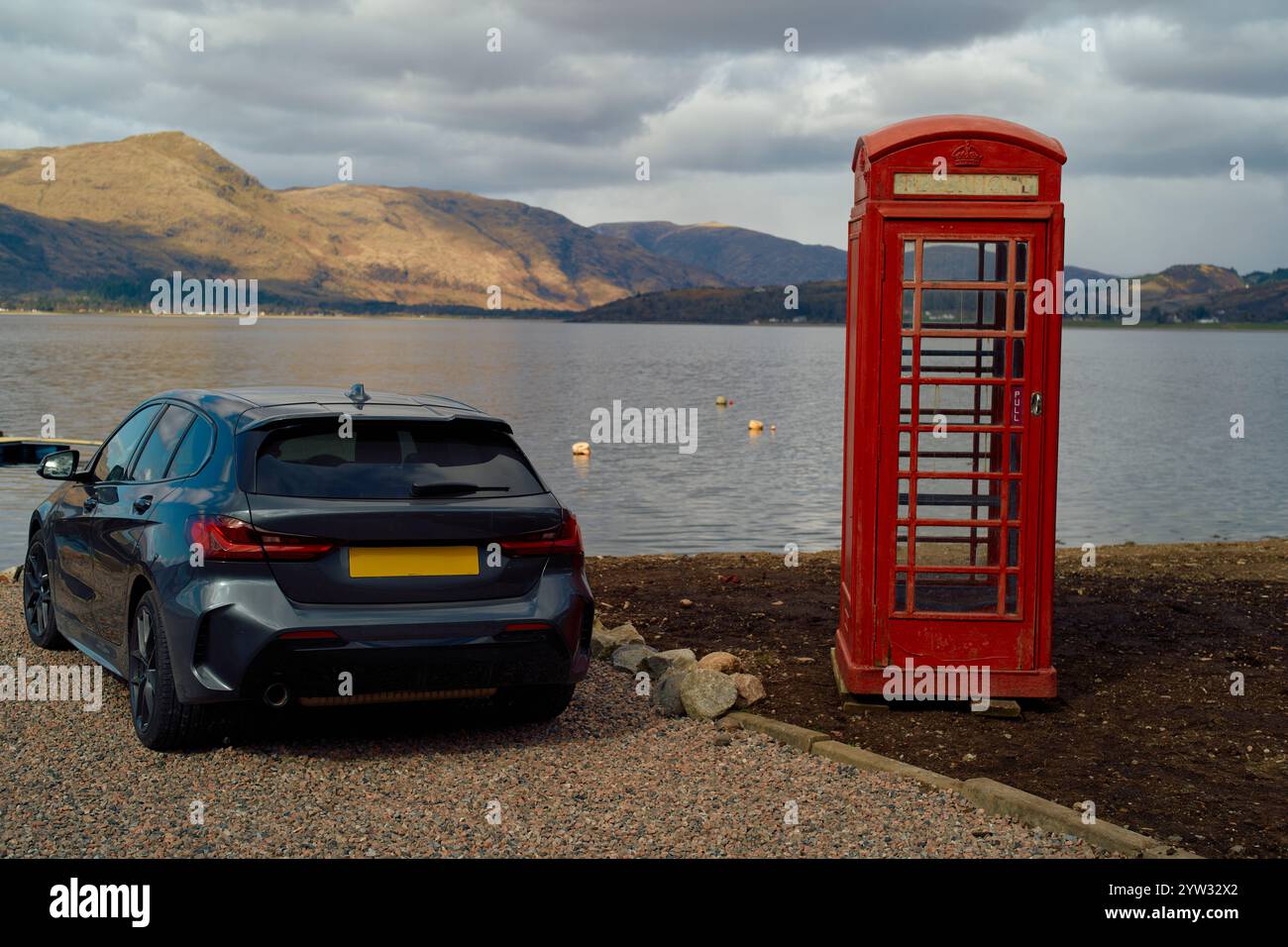 A red phone booth stands beside a dark blue car with a scenic lake and ...