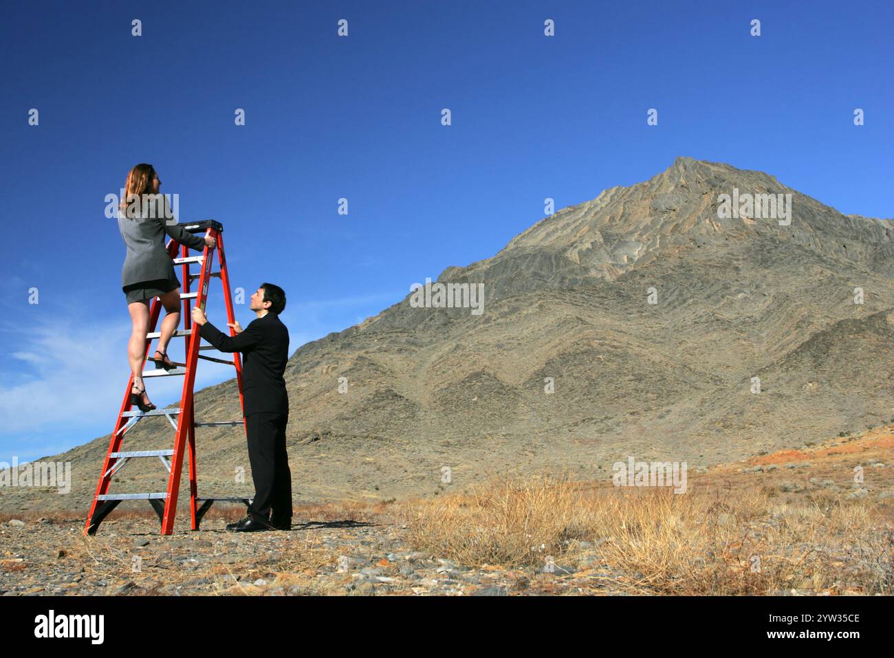 A woman steps up on a ladder in front of a mountain while her colleague ...