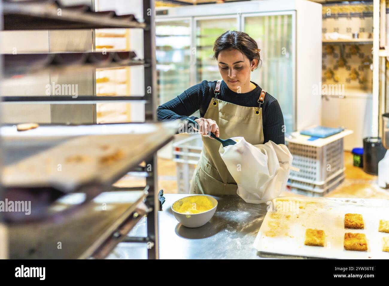 Female latin artisan baker filling a piping bag to decorate pastries in the workshop kitchen of ...