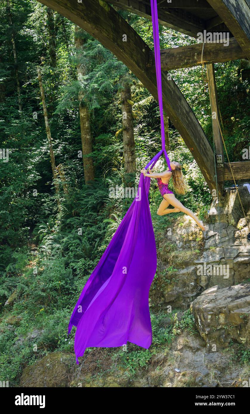 Aerial performer poses with purple fabric under a bridge in a lush ...