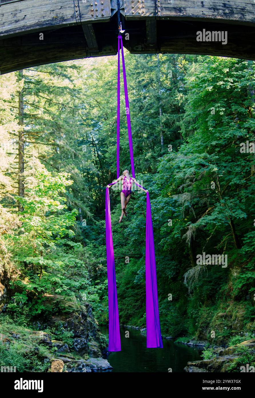Aerial silk performer executing a pose under a wooden bridge in a lush green forest setting, USA ...