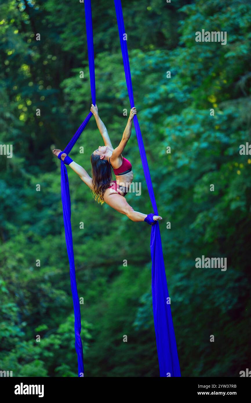 Aerial silk performer executing a pose in bright blue fabric against a lush green forest ...