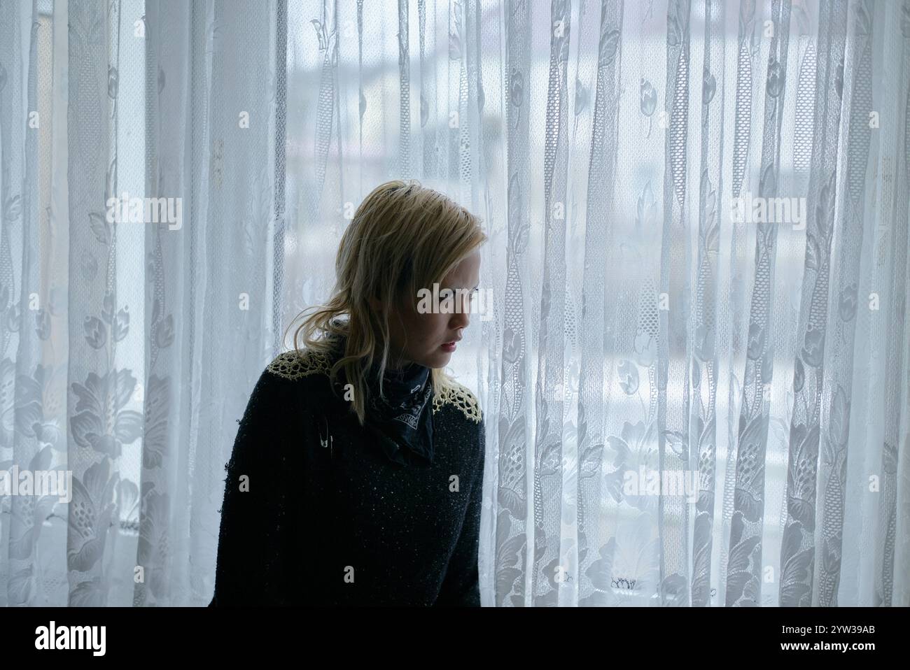 A contemplative adult woman stands by a window, looking outside through ...