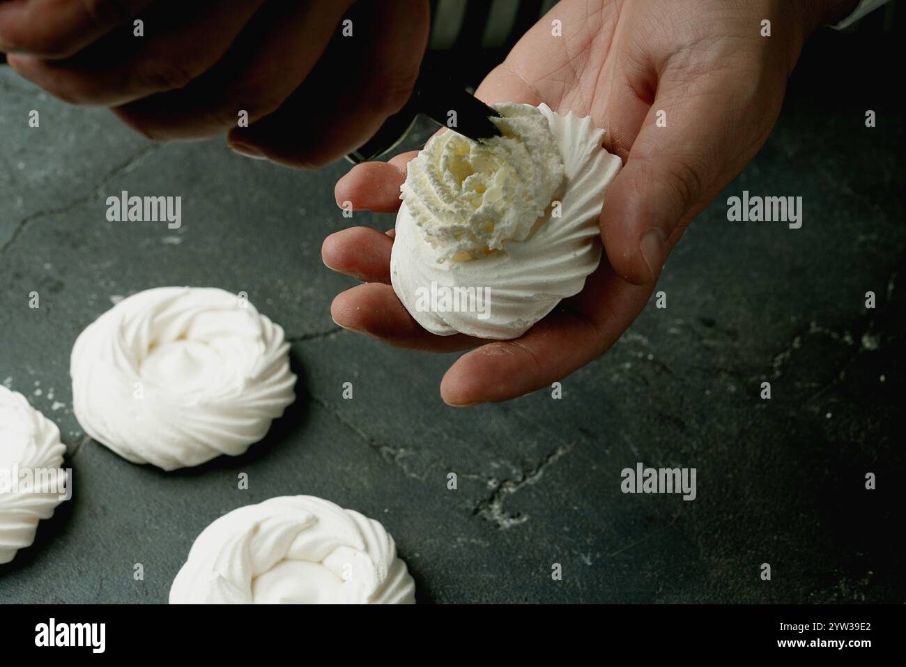 Hands piping whipped cream onto meringue nests. Dark textured surface ...