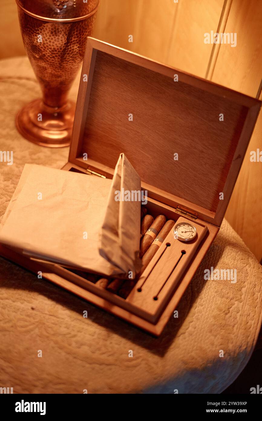 Wooden cigar box open on a fabric surface with cigars and a hygrometer inside, illuminated by ...