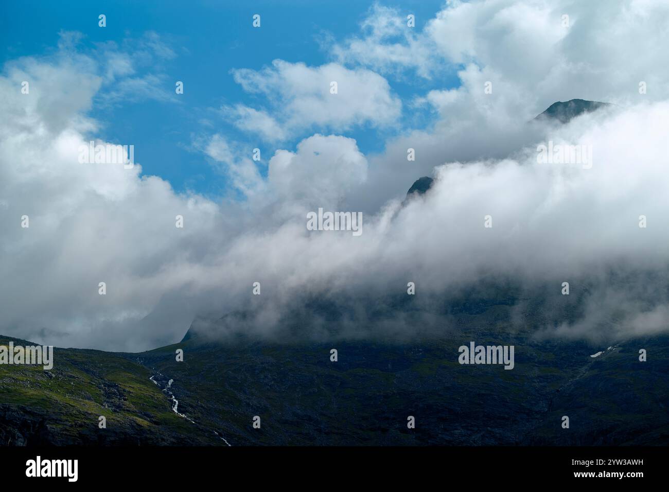 Clouds shroud rugged mountain peaks under a blue sky with sunlight ...