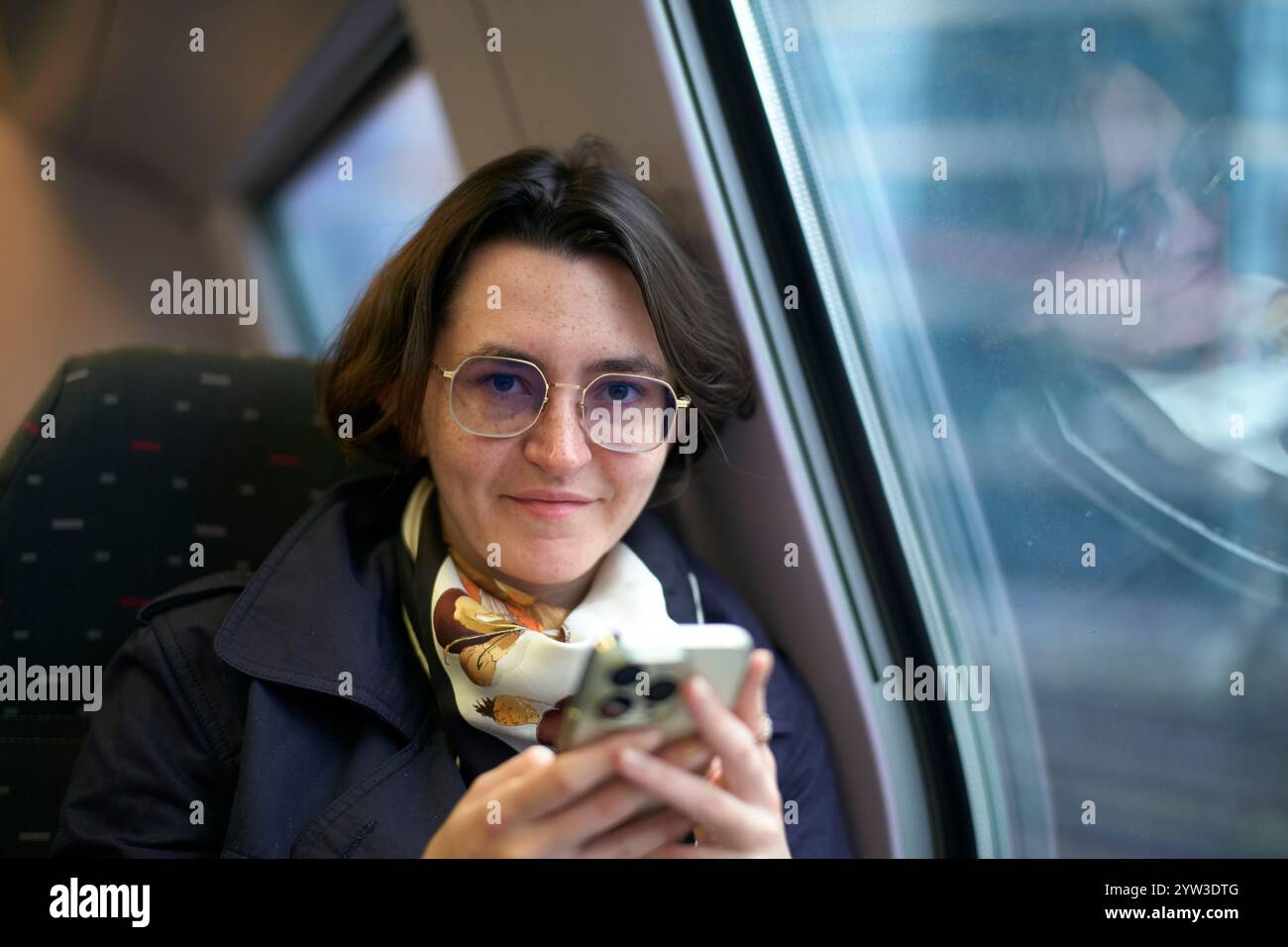 Woman with glasses using her smartphone on a train, with her reflection ...