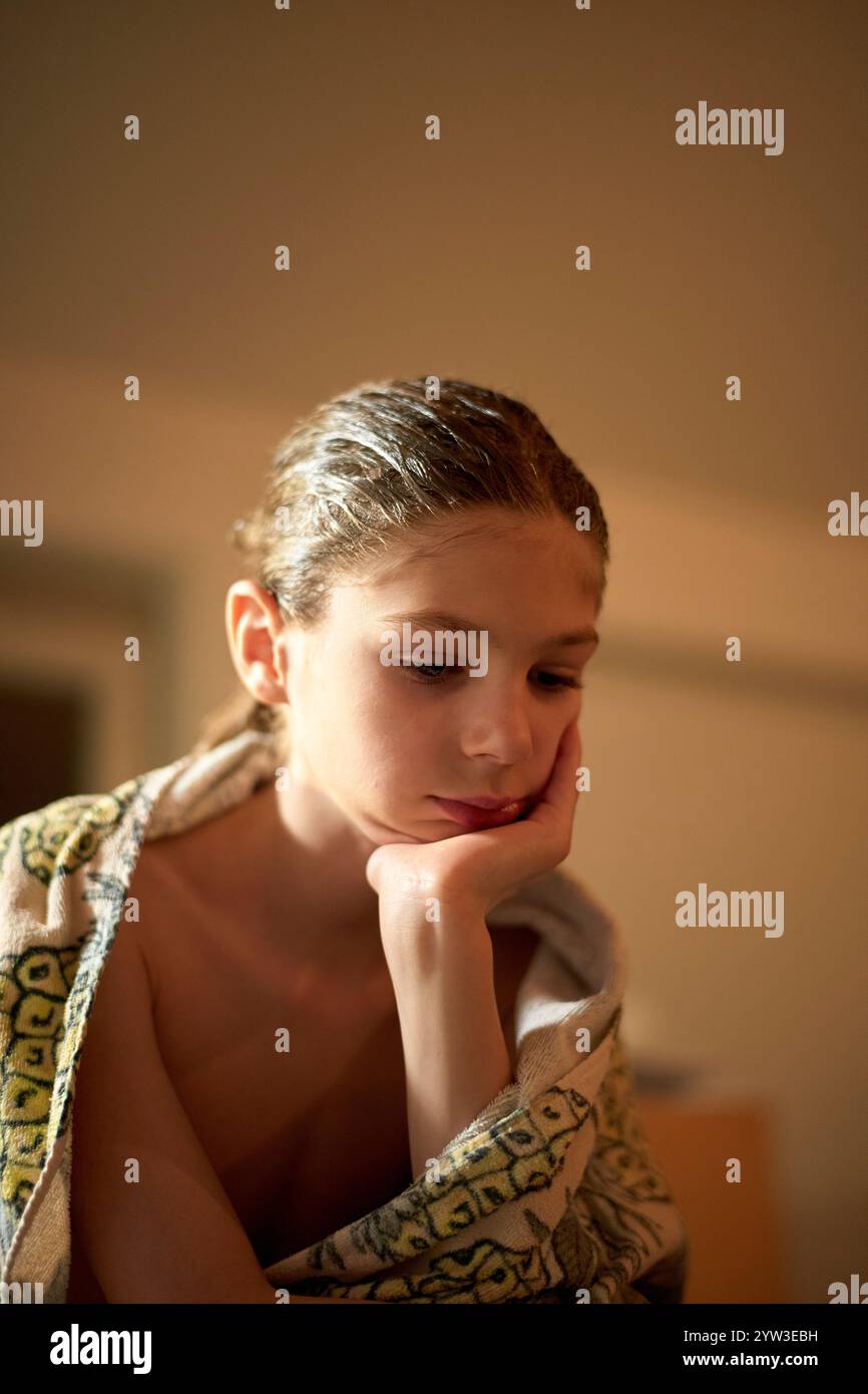 A contemplative young boy with wet hair wrapped in a towel rests his ...
